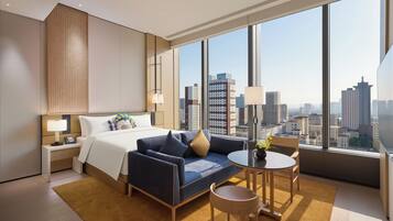 Quarto de hotel moderno com cama grande, sofá, mesinha e vista para a paisagem urbana.