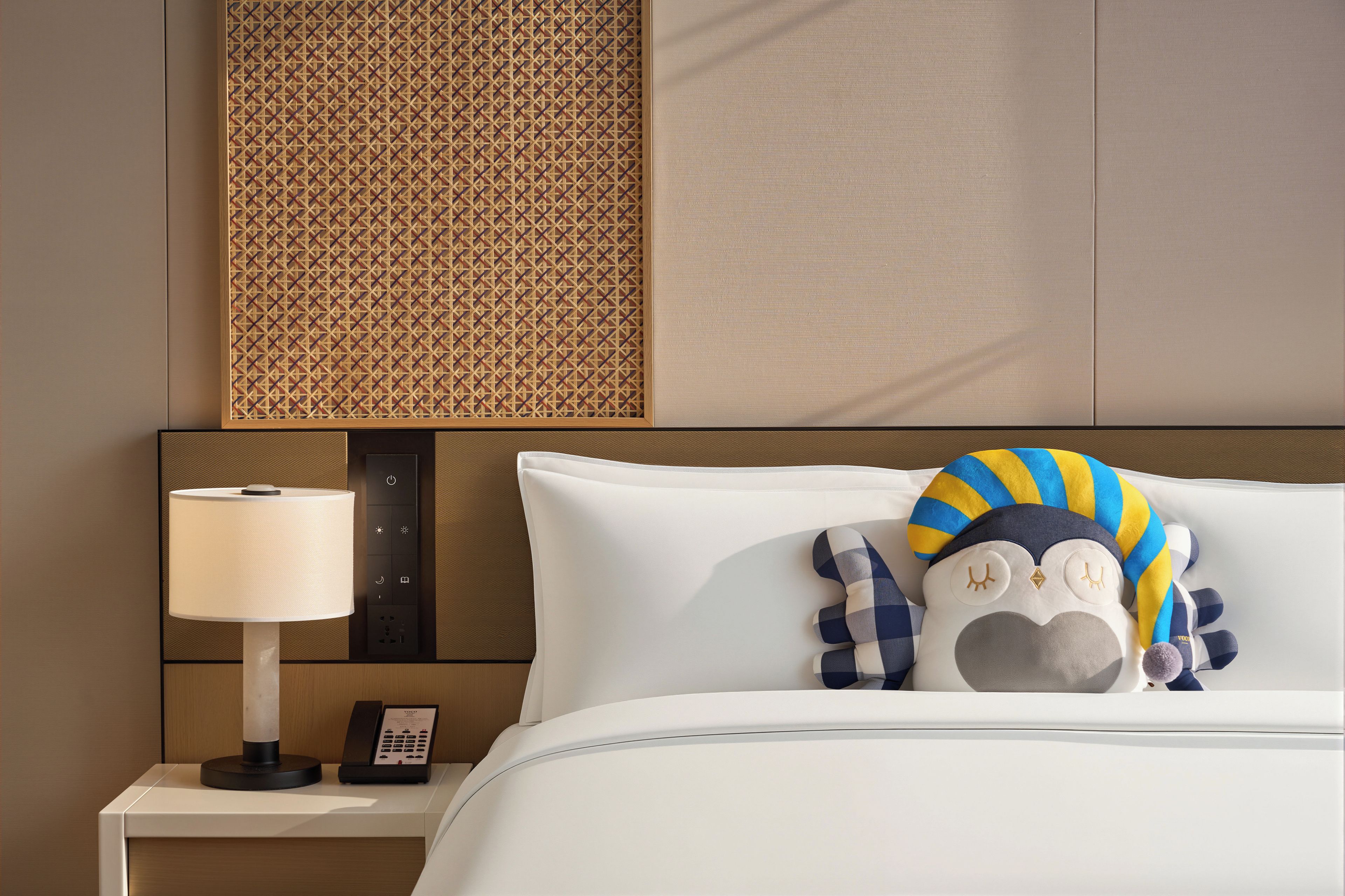 Quarto de hotel com cama, mesa de cabeceira com abajur, telefone e almofada decorativa.