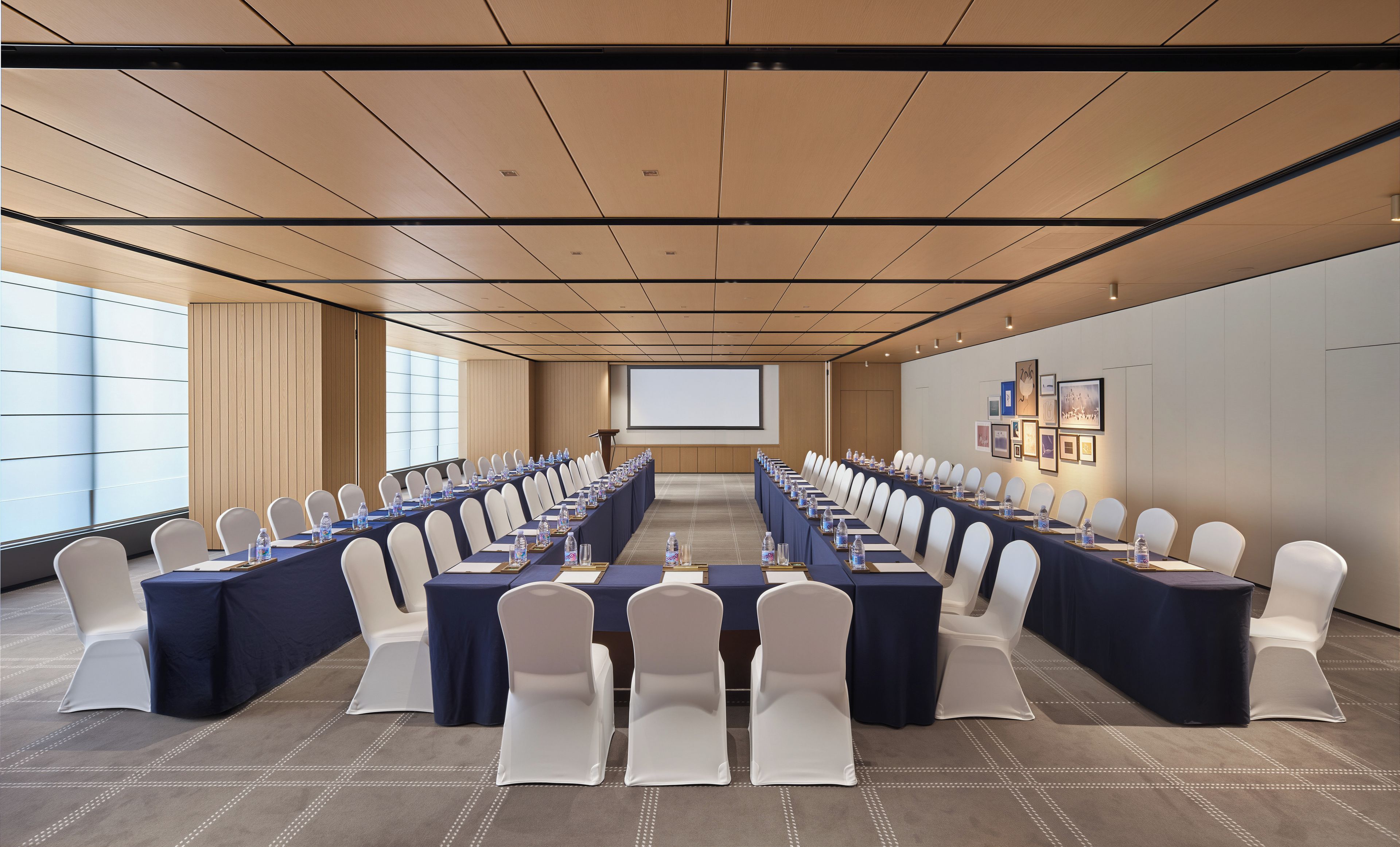 Uma sala de conferências com uma mesa comprida, cadeiras brancas e uma tela de projeção.