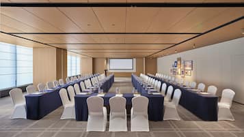 Uma sala de conferências com uma mesa comprida, cadeiras brancas e uma tela de projeção.