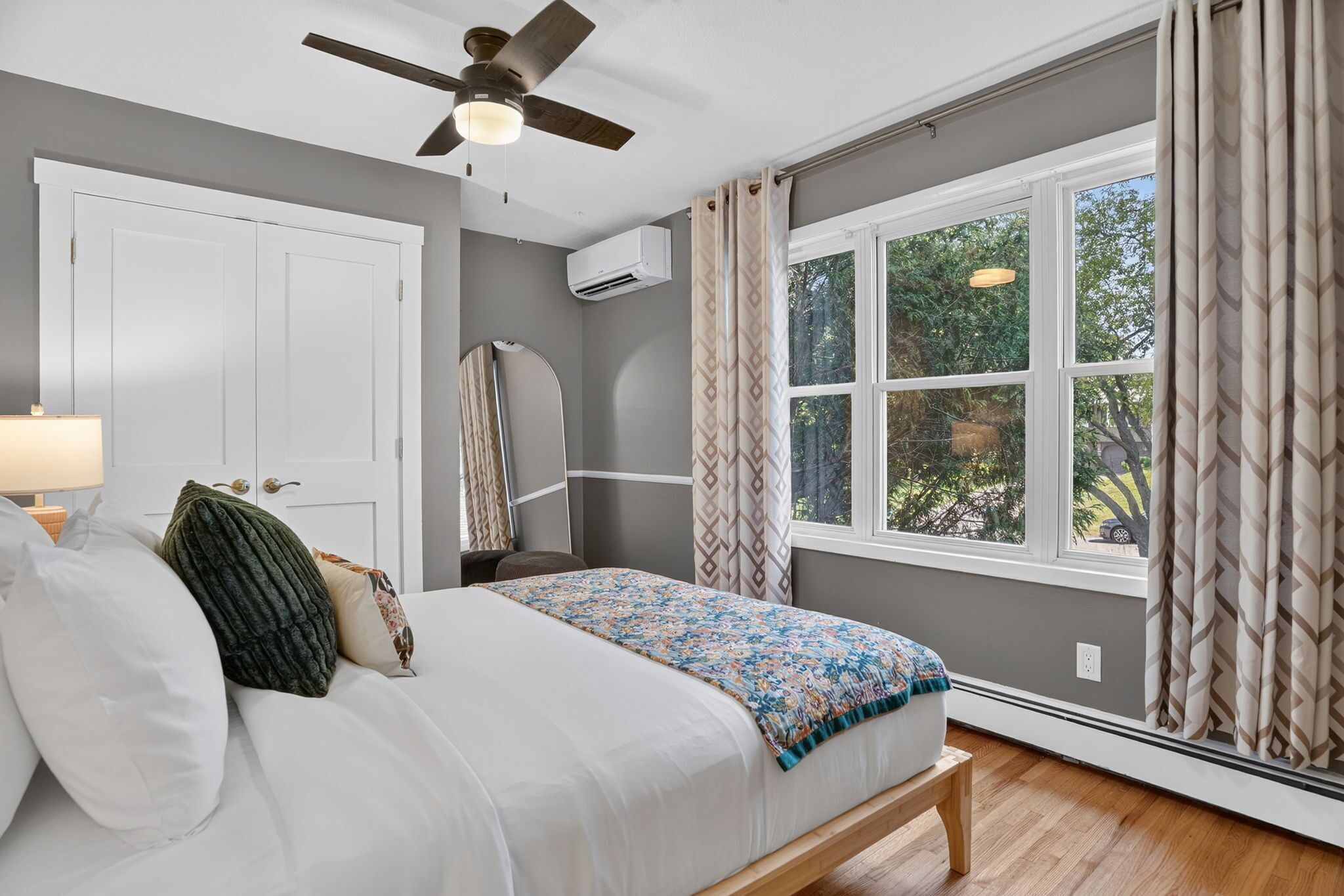 Quarto com cama, ventilador de teto, ar condicionado, janela com cortinas e vista para o verde lá fora.