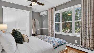 Quarto com cama, ventilador de teto, ar condicionado, janela com cortinas e vista para o verde lá fora.