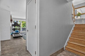 Um corredor com escadas de madeira que levam a um patamar, uma porta e vista para uma área de ginástica com equipamentos.
