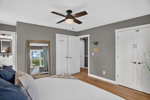 Quarto com cama, ventilador de teto, espelho grande, guarda-roupa e porta para outro cômodo.