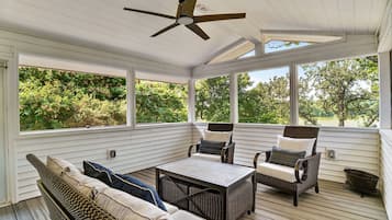 Varanda coberta com móveis de vime, ventilador de teto e grandes janelas com vista para uma área arborizada.