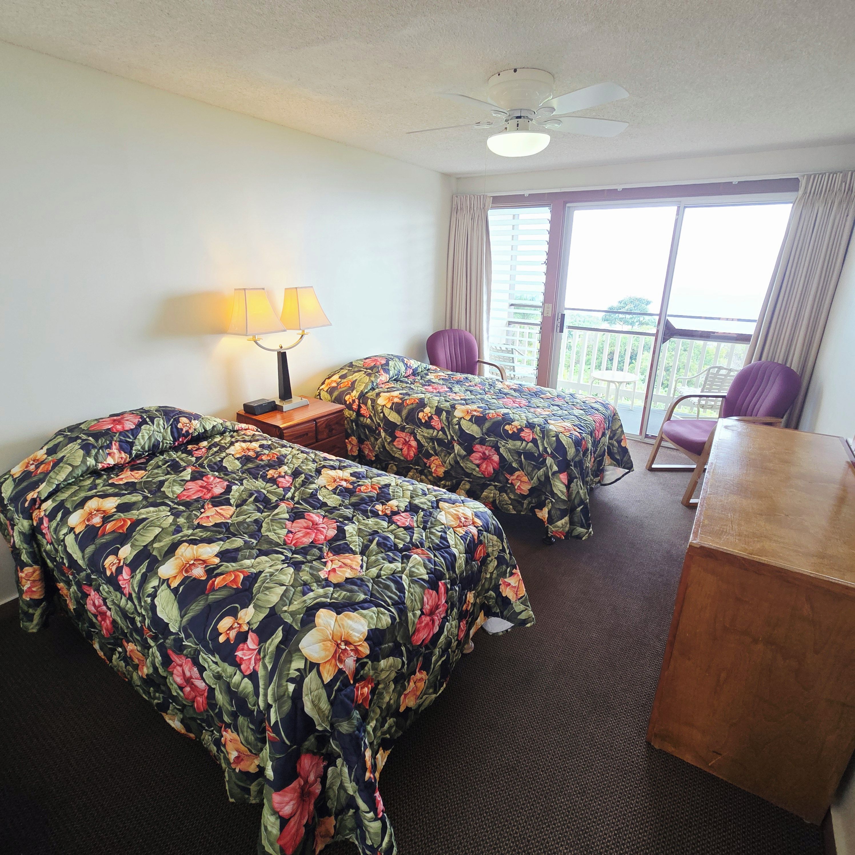 Quarto de hotel com duas camas, uma escrivaninha, uma cadeira, um ventilador de teto e uma janela com cortinas.