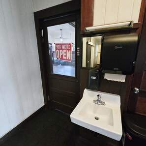 Um banheiro com uma pia branca, um espelho e uma porta com a placa 'YES WE'RE OPEN'.