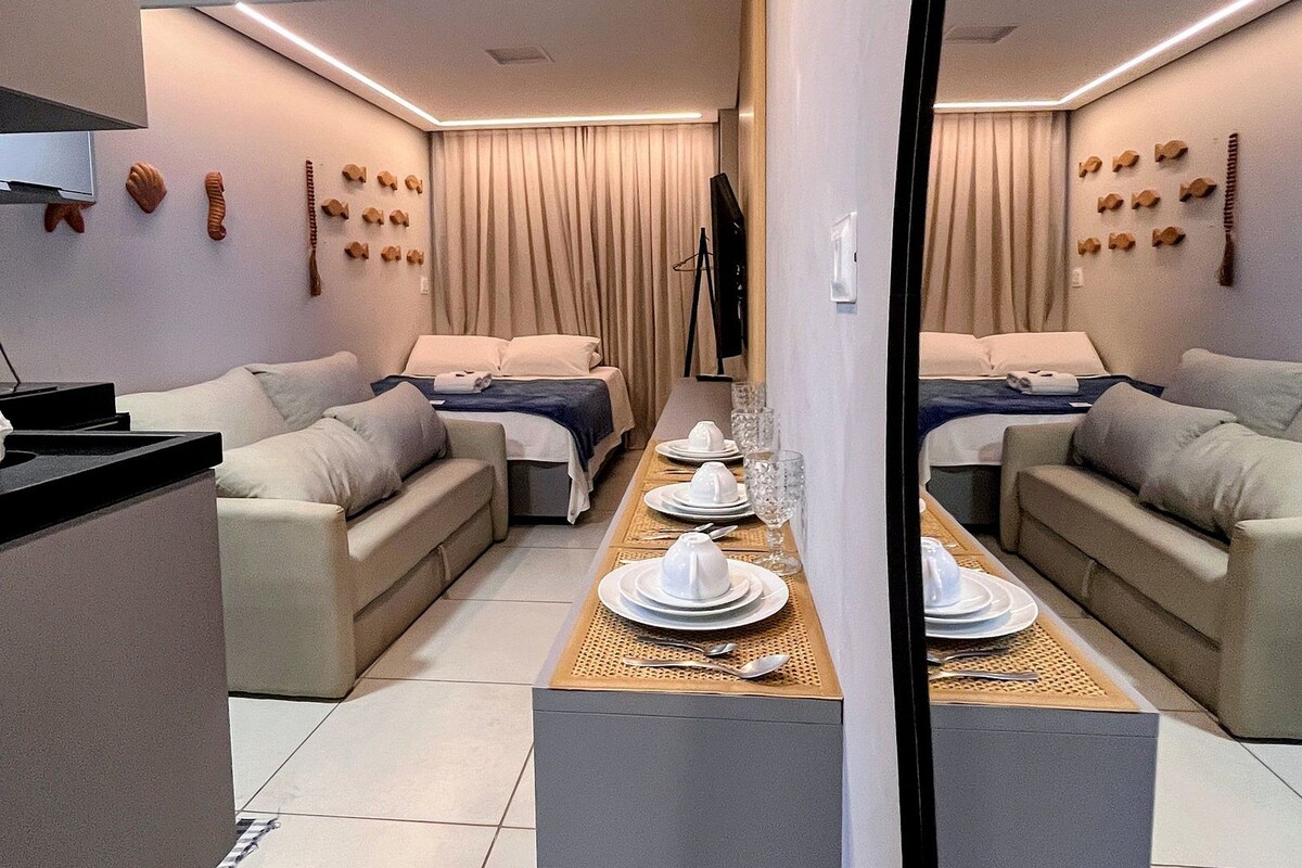 Quarto de hotel moderno com área de jantar, dois sofás, TV e minibar.