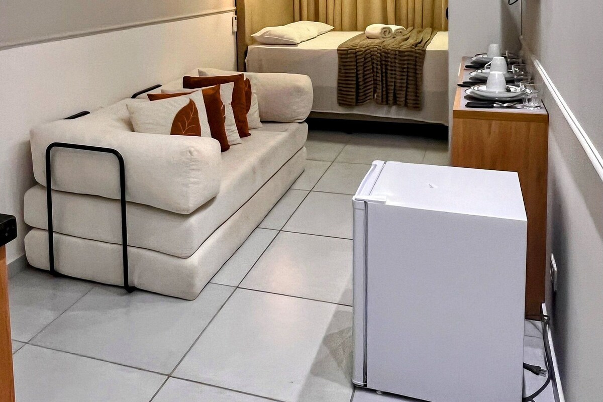 Quarto de hotel compacto com cama, sofá, pequeno frigorífico, televisão e obras de arte na parede.