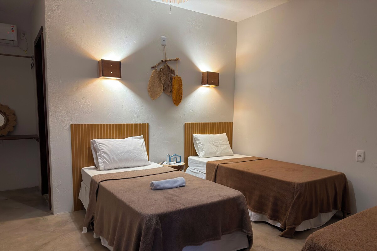 Quarto com duas camas, cada uma com colcha marrom e almofadas brancas, uma pequena mesa de cabeceira com abajur, e luminárias fixadas na parede.
