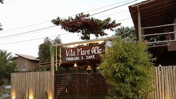 Uma entrada rústica com um portão de madeira, uma placa indicando Vila Mare Alta, e uma trilha de terra que leva a um edifício com telhado inclinado.