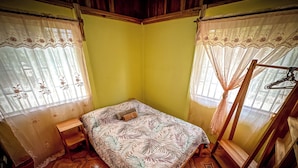 Quarto pequeno com uma cama de solteiro, colcha estampada, um banco de madeira e mosquiteiro.