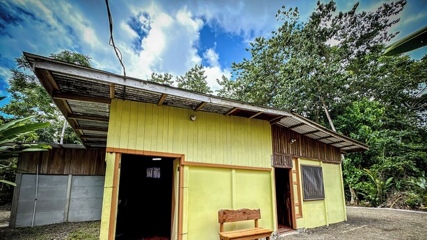 Um pequeno edifício térreo com telhado inclinado, um banco na frente e uma porta que dá para o interior.