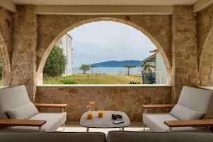 Quarto com paredes de pedra e vista para um corpo de água e montanhas. Há dois sofás e uma mesa pequena com um copo de suco de laranja e um smartphone.