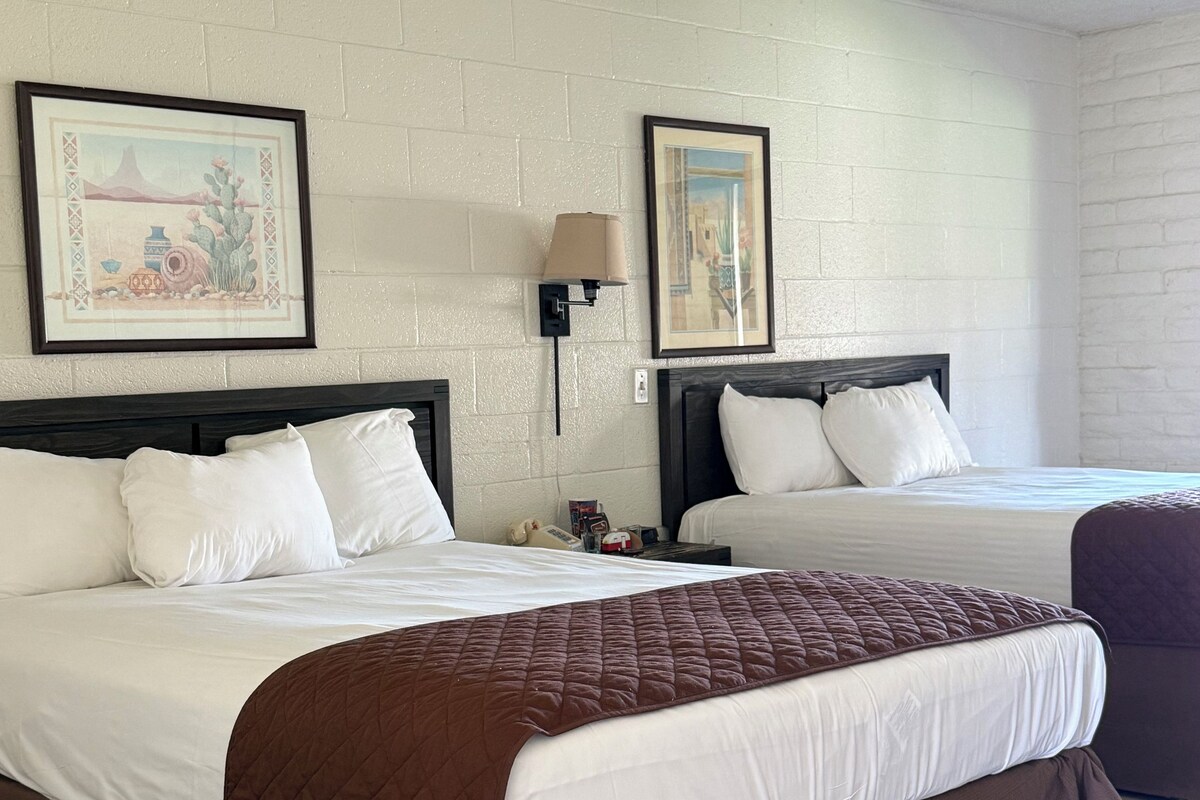 Quarto de hotel com duas camas, parede de tijolos brancos, quadro emoldurado, luminária na parede e piso de azulejos.