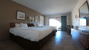 Quarto de hotel com duas camas, uma escrivaninha, uma cadeira, televisão e porta de correr para uma varanda.