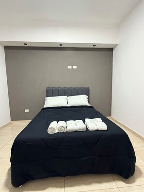 Cama bem arrumada com edredom preto e almofadas brancas, acompanhada de toalhas brancas enroladas.