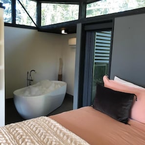 Quarto moderno com uma banheira grande, cama com travesseiro rosa e preto, e vista pela janela.