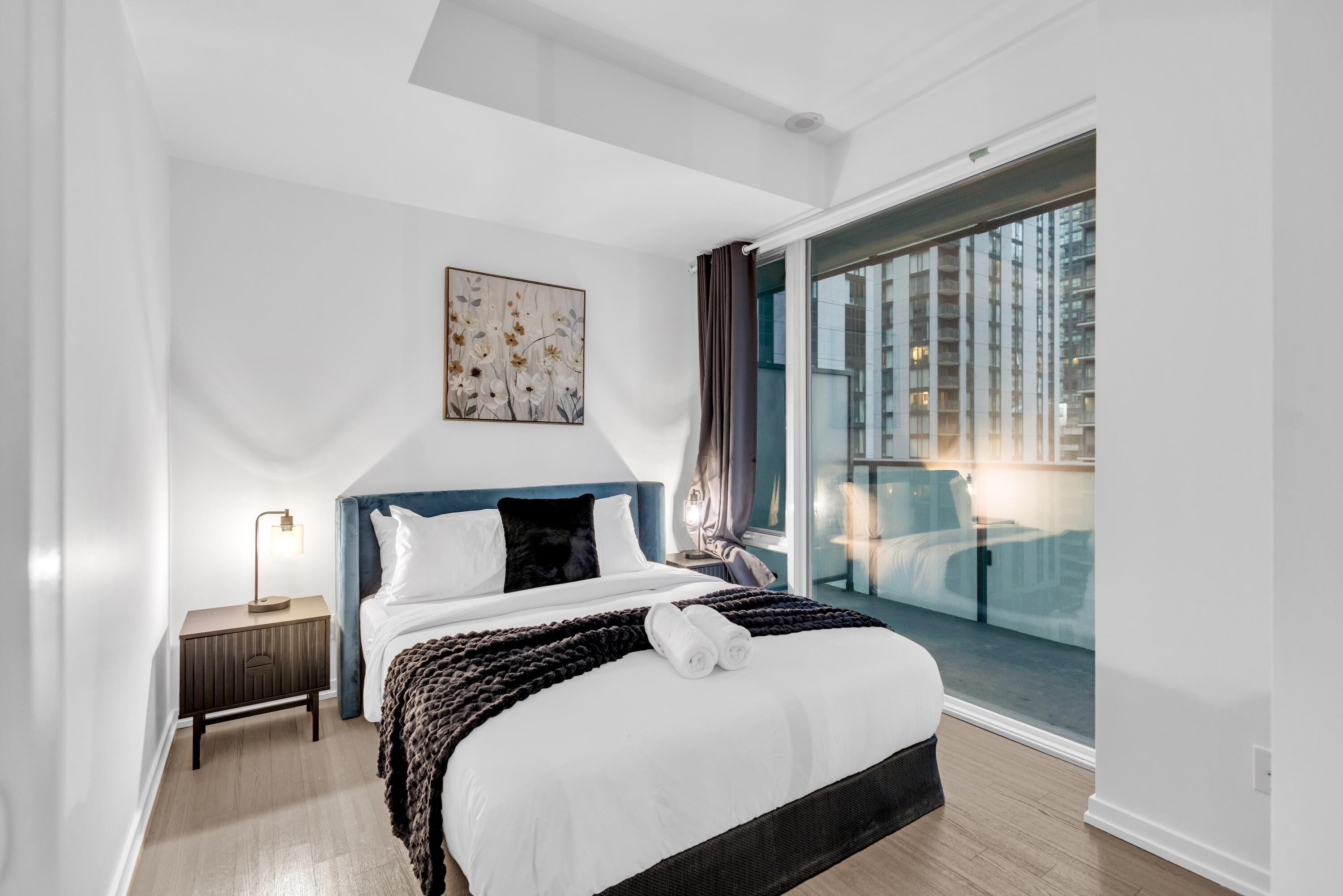 Quarto de hotel moderno com uma cama grande, uma mesa de cabeceira, uma luminária e uma janela com vista para edifícios.