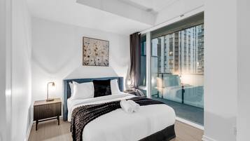 Quarto de hotel moderno com uma cama grande, uma mesa de cabeceira, uma luminária e uma janela com vista para edifícios.