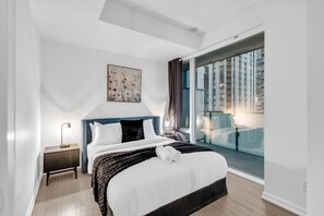 Quarto de hotel moderno com uma cama grande, uma mesa de cabeceira, uma luminária e uma janela com vista para edifícios.