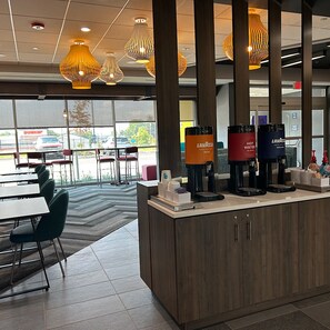 Estação de café moderna com diversos dispensadores, incluindo água quente e opções regulares, em espaço amplo com assentos e janelas grandes.
