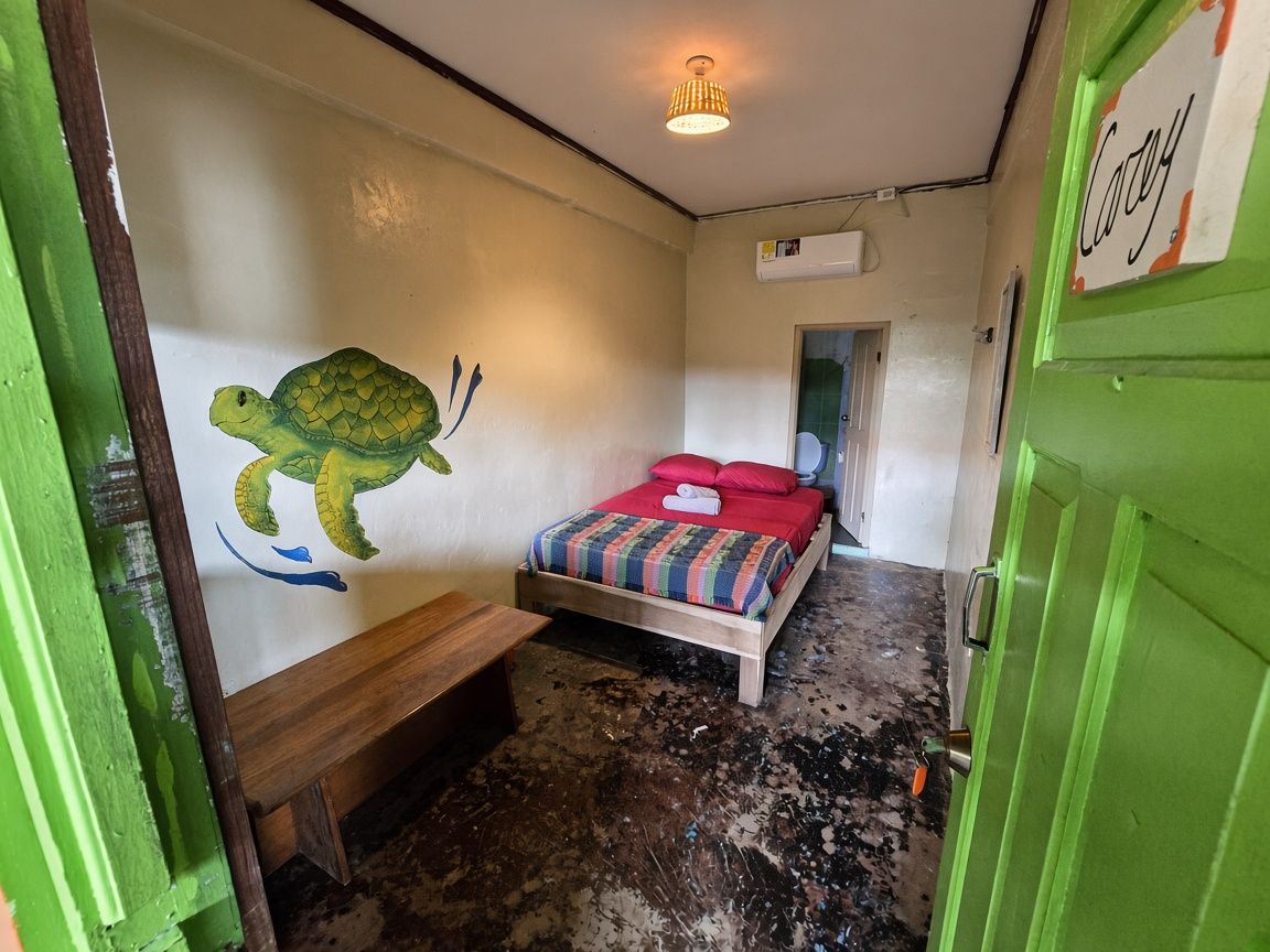 Quarto com cama, um mural na parede, um banco e uma porta com um aviso.