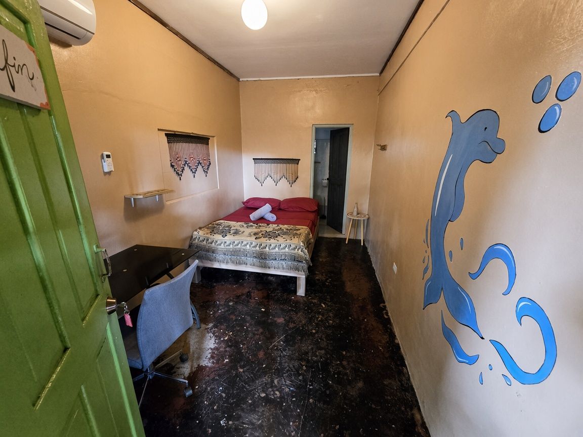 Quarto pequeno com uma cama, escrivaninha e cadeira. Há uma decoração na parede e uma porta.