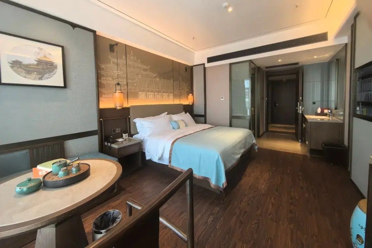 Quarto de hotel com uma cama, mesa de cabeceira e escrivaninha com uma cadeira. Há uma obra de arte na parede e uma janela.