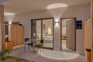 Quarto de hotel moderno com porta de vidro deslizante que dá para o quarto, uma pequena mesa com cadeiras e uma placa indicando o número do quarto S.02.