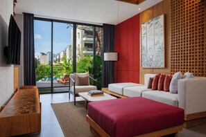 Quarto de hotel moderno com uma cama grande, um sofá, uma mesa de centro e uma TV. Há uma varanda com piscina e vegetação visíveis lá fora.