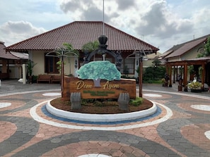 Uma praça circular com uma placa de madeira central com a inscrição 'Dem Ayem', rodeada por um pavimento estampado.