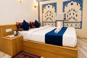Quarto de hotel com uma cama, uma mesa de cabeceira com uma chaleira e um tabuleiro com chávenas, além de um tapete vermelho.