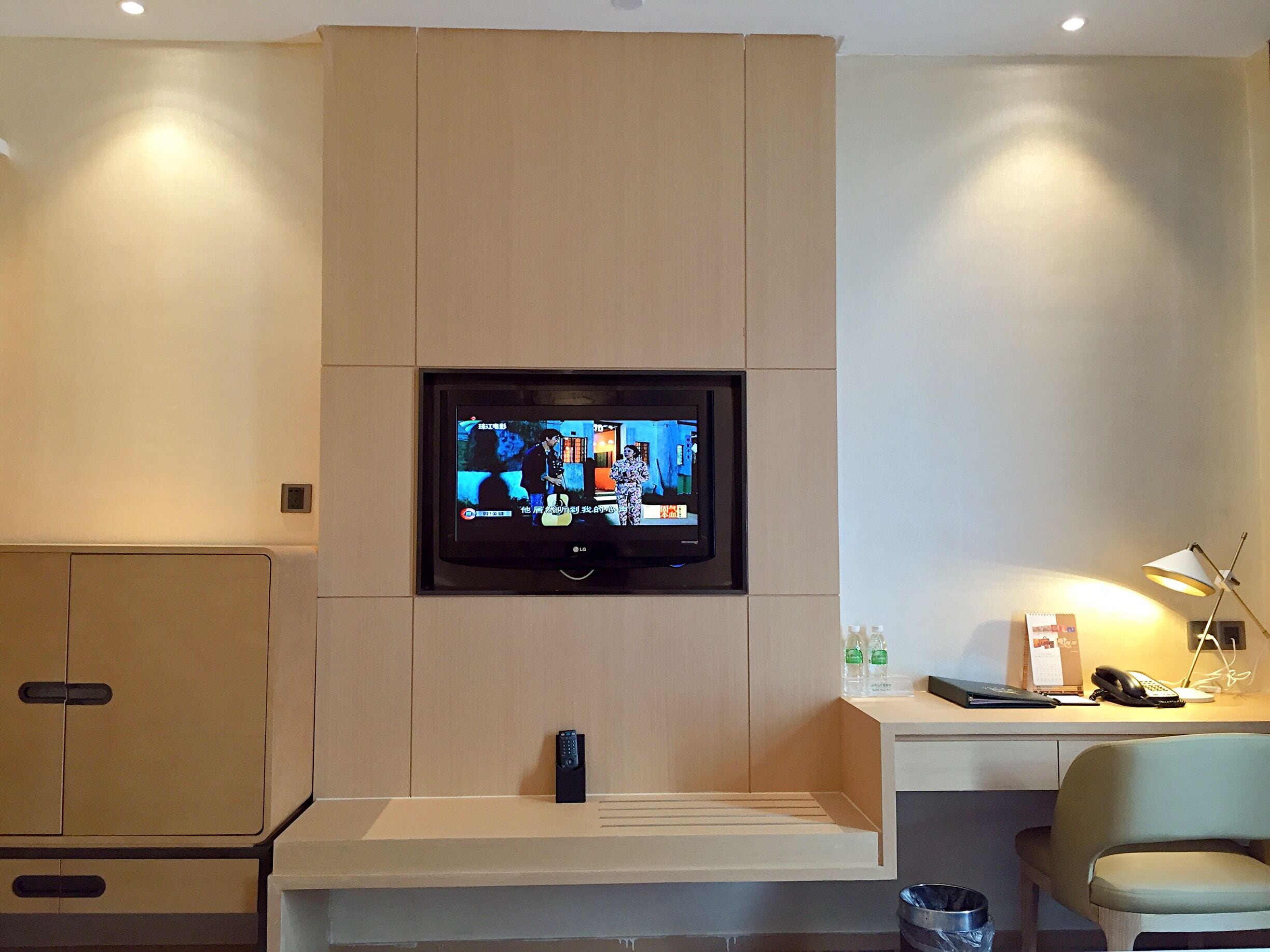 Quarto de hotel com televisão montada, uma mesa com luminária e uma cadeira.