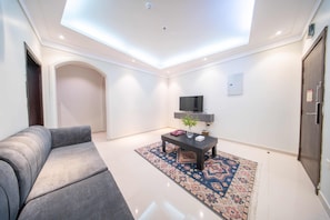 Uma sala de estar moderna com um sofá cinzento, uma televisão de tela plana sobre um suporte, uma mesa de centro com objetos decorativos e um tapete estampado.