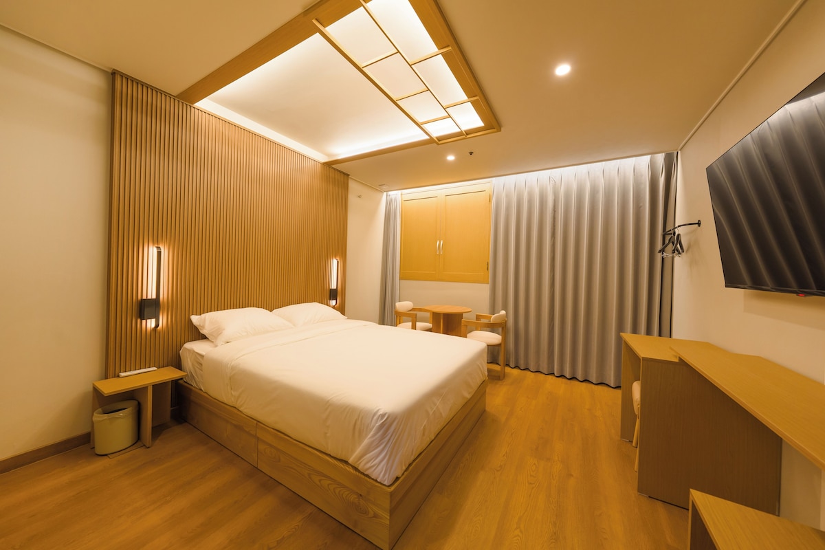 Quarto de hotel moderno com cama de madeira, escrivaninha, cadeira, televisão e uma grande janela com cortinas.