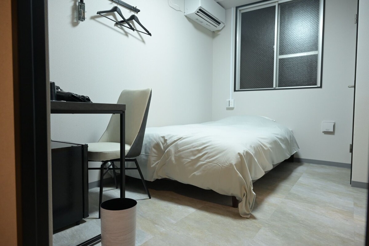 Quarto pequeno e limpo, com cama, cadeira, escrivaninha e janela.