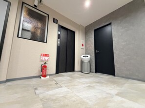 Corredor com extintor, lixeira e porta com luz de emergência vermelha.
