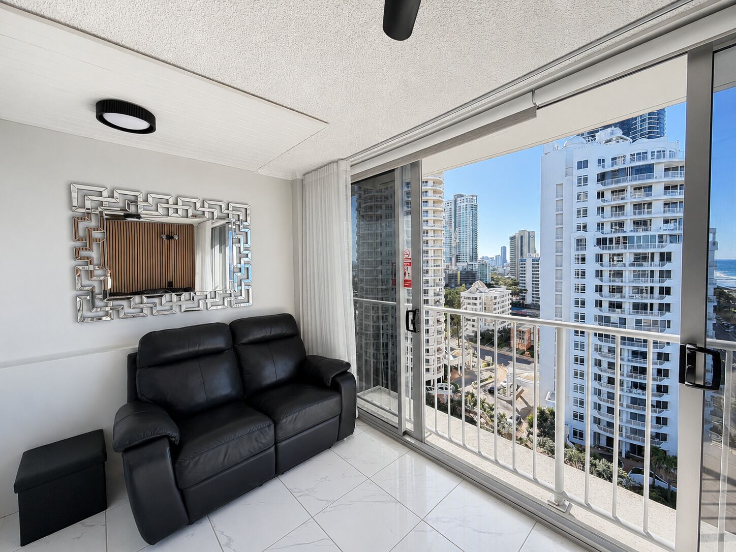 Uma sala de estar moderna com um sofá de couro preto, um espelho decorativo e vista para a cidade através de grandes janelas.