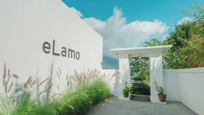 Um edifício branco com o texto 'eLamo' na fachada, um caminho de concreto que leva a um arco branco e uma pequena área de jardim com plantas em vasos.