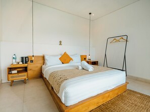 Quarto com cama de madeira, roupa de cama branca, mesa de cabeceira de madeira, uma garrafa de água, chaleira e um cabideiro.