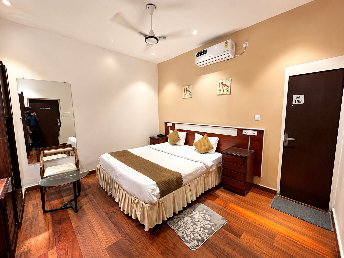 Quarto de hotel com cama, mesas de cabeceira, espelho, ventoinha no teto e ar condicionado, além de uma porta com o número do quarto.