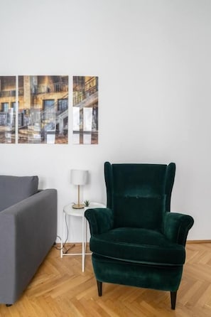 Um quarto com uma poltrona de veludo verde, um sofá cinzento e uma mesa de apoio branca com uma luminária.