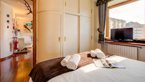 Quarto de hotel com cama, televisão, janela com cortinas e vista para edifícios.