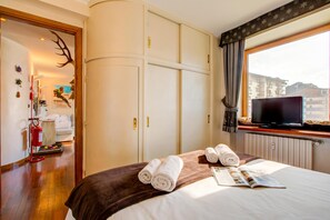 Quarto de hotel com cama, televisão, janela com cortinas e vista para edifícios.