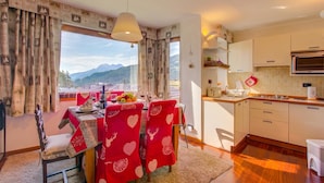 Uma área de refeições com uma mesa posta para dois, uma cozinha equipada com micro-ondas e vista para montanhas através de grandes janelas.