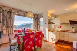 Uma área de refeições com uma mesa posta para dois, uma cozinha equipada com micro-ondas e vista para montanhas através de grandes janelas.