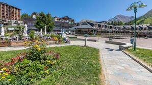 Uma praça de cidade com uma fonte central, rodeada por edifícios com varandas e uma montanha ao fundo.