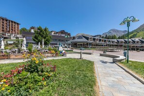 Uma praça de cidade com uma fonte central, rodeada por edifícios com varandas e uma montanha ao fundo.