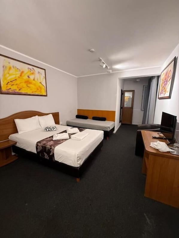 Quarto de hotel com cama, escrivaninha, cadeira e porta que dá para outro cômodo.